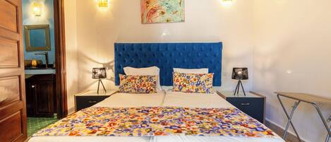 Quarto com cama grande, duas mesas de cabeceira, cabeceira estofada azul e uma pintura abstrata colorida sobre a cama.