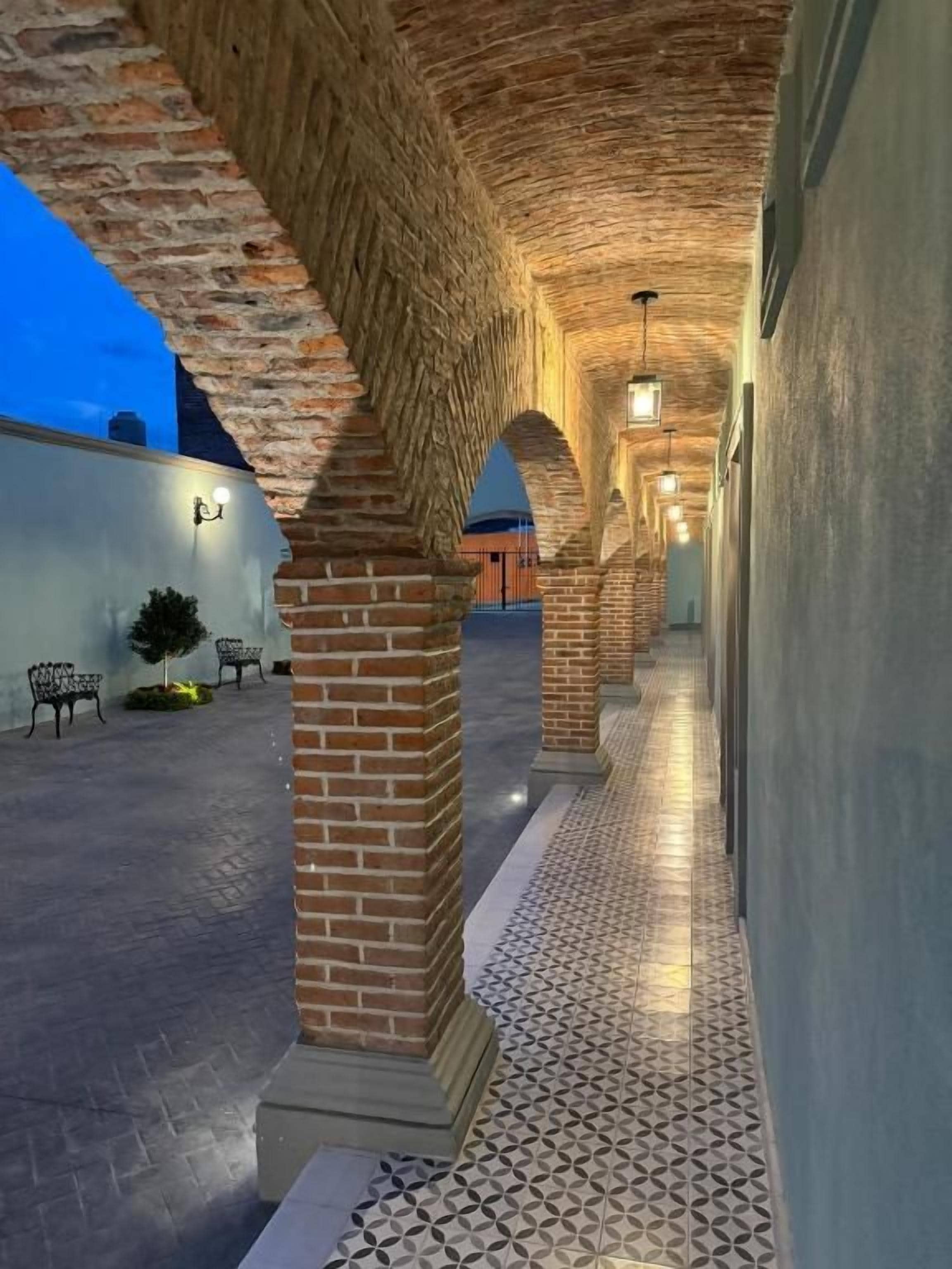 Um corredor com arcos de tijolo e piso estampado, conduzindo a um edifício com telhado plano.