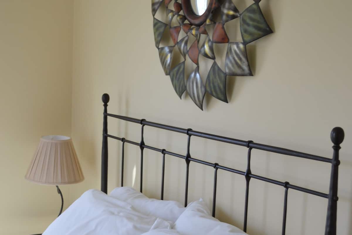 Uma cama bem arrumada com lençóis brancos, cabeceira de metal e um espelho decorativo fixado na parede.
