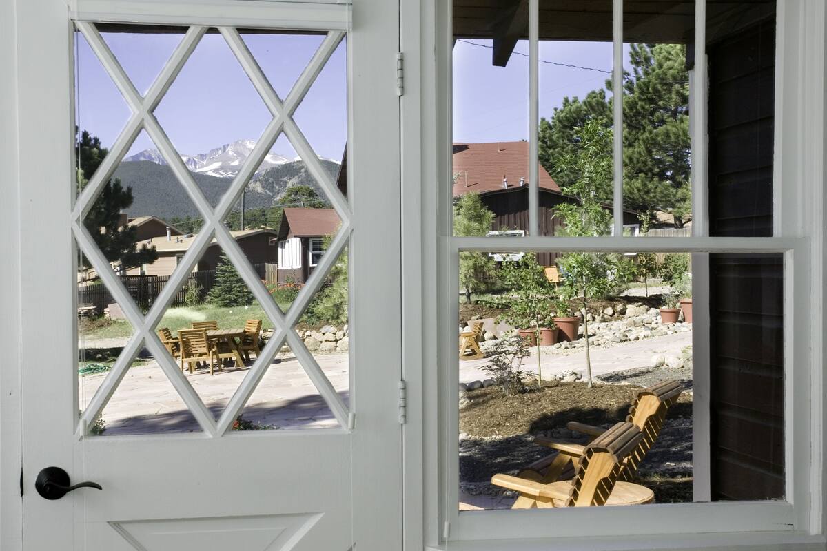 Vista pela janela com moldura branca, mostrando um espaço de pátio com cadeiras e mesa de madeira, um jardim com plantas em vasos e uma cordilheira ao fundo.