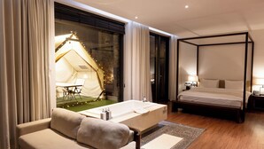 Quarto de hotel moderno com uma cama grande, um sofá, uma banheira e uma área externa coberta visível pela janela.