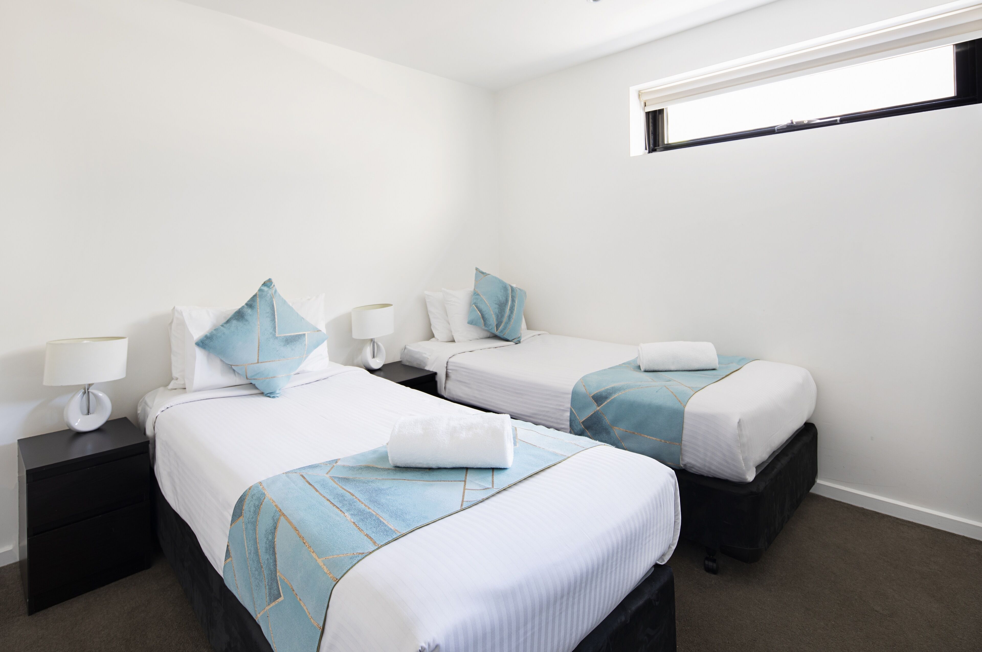 Quarto de hotel com duas camas, cada uma com colcha branca e azul, uma mesa de cabeceira com abajur e uma janela com persianas.