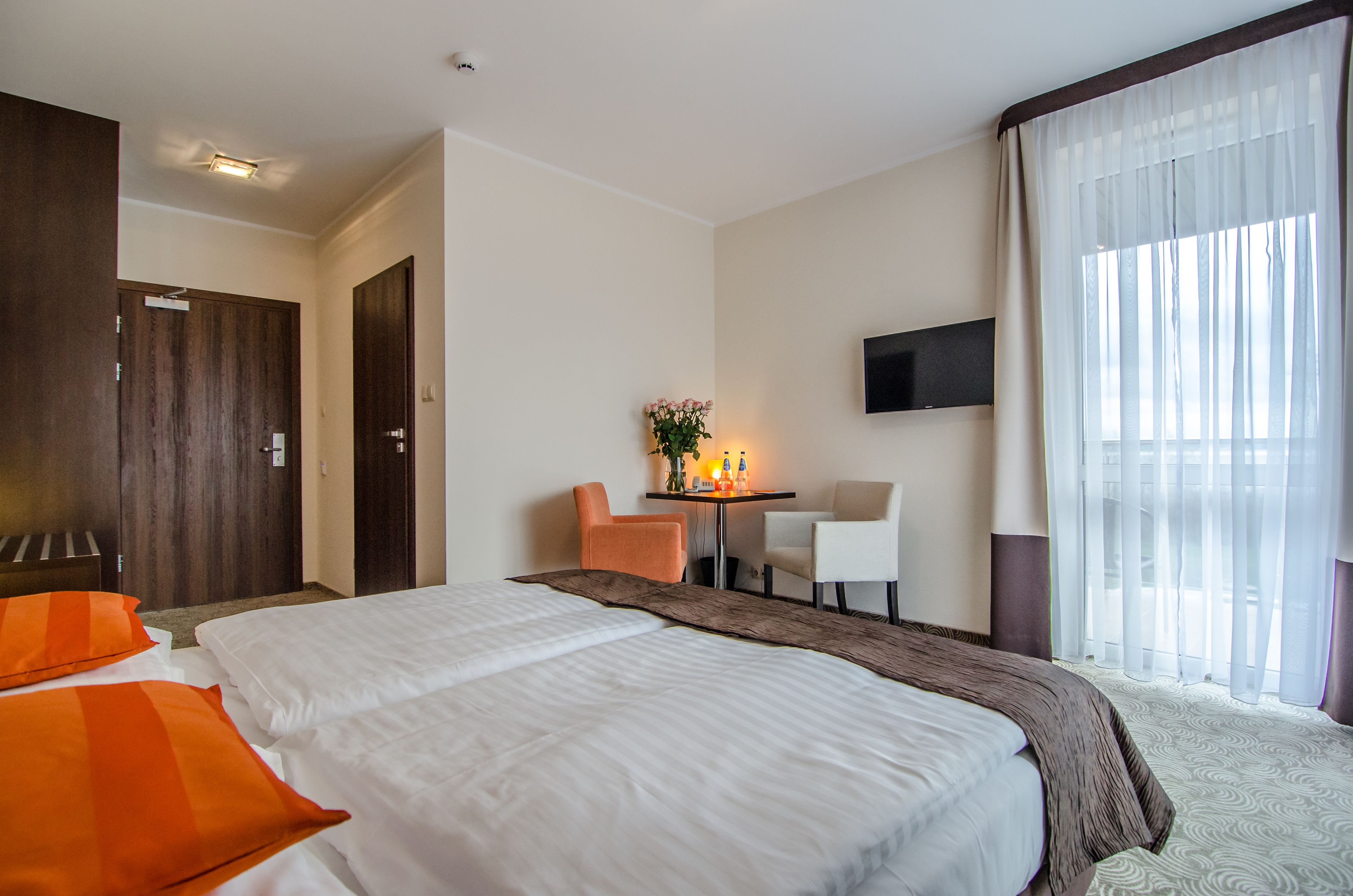 Quarto de hotel com uma cama grande, duas cadeiras, uma escrivaninha, televisão e janela com cortinas.