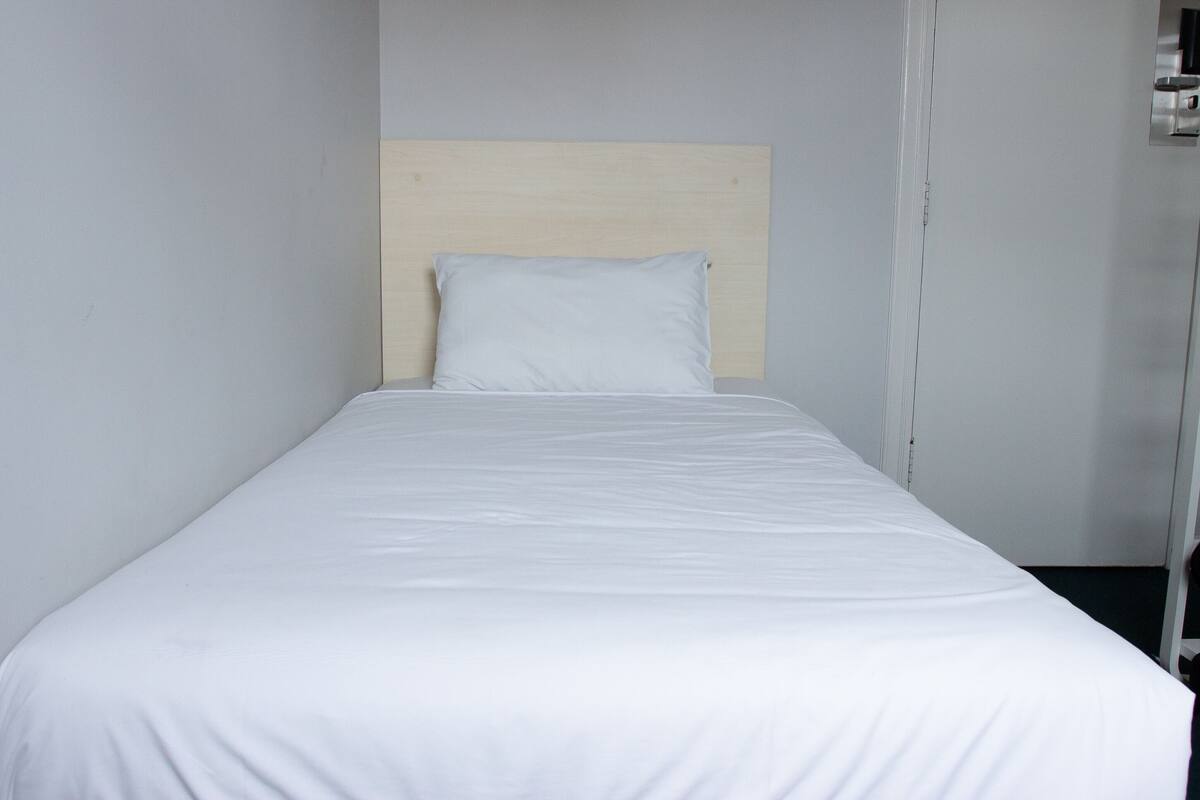 Cama bem arrumada com cabeceira de madeira e roupa de cama branca em um quarto simples com paredes em tons claros.
