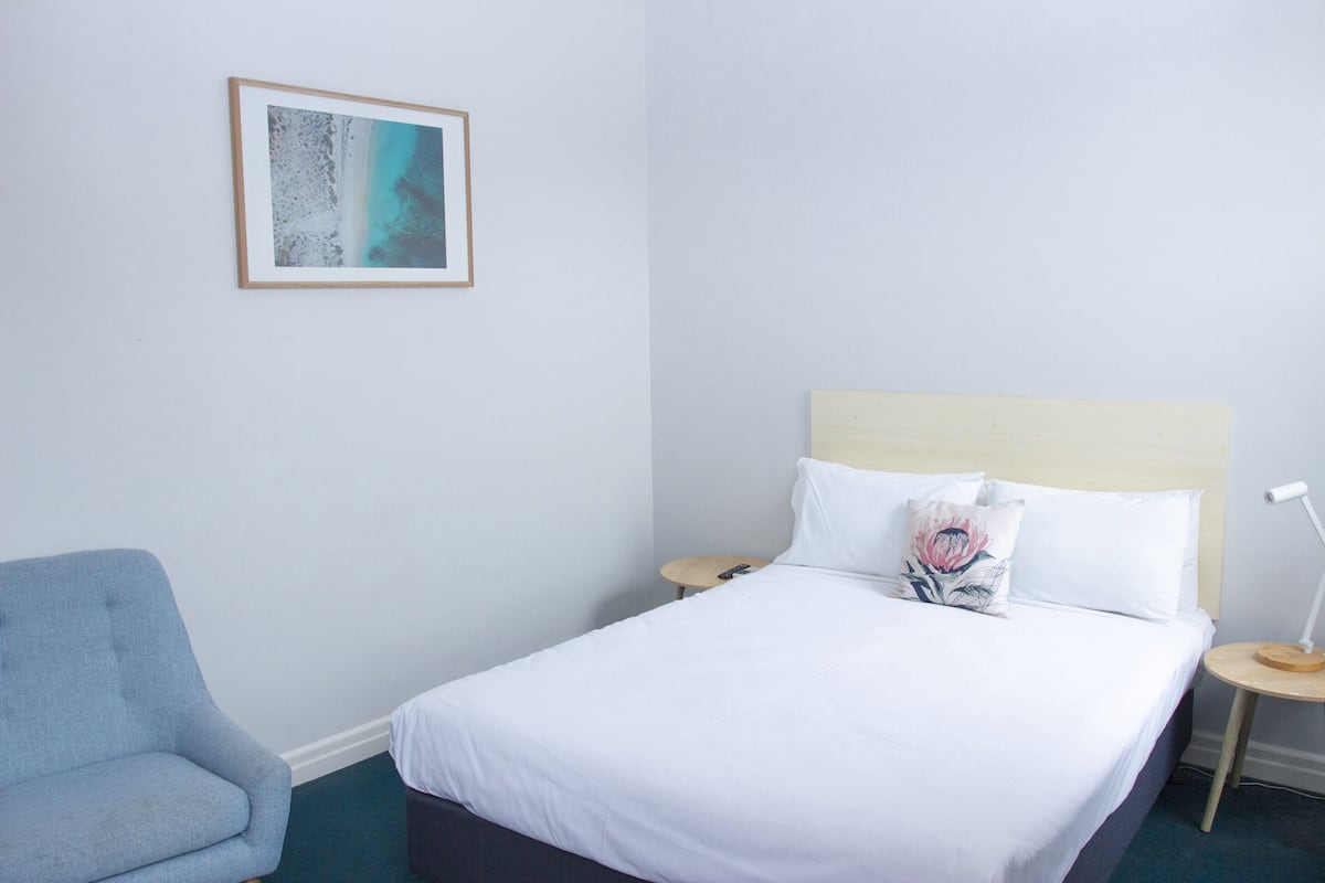 Quarto de hotel com cama, cadeira, mesa de cabeceira e um quadro na parede.