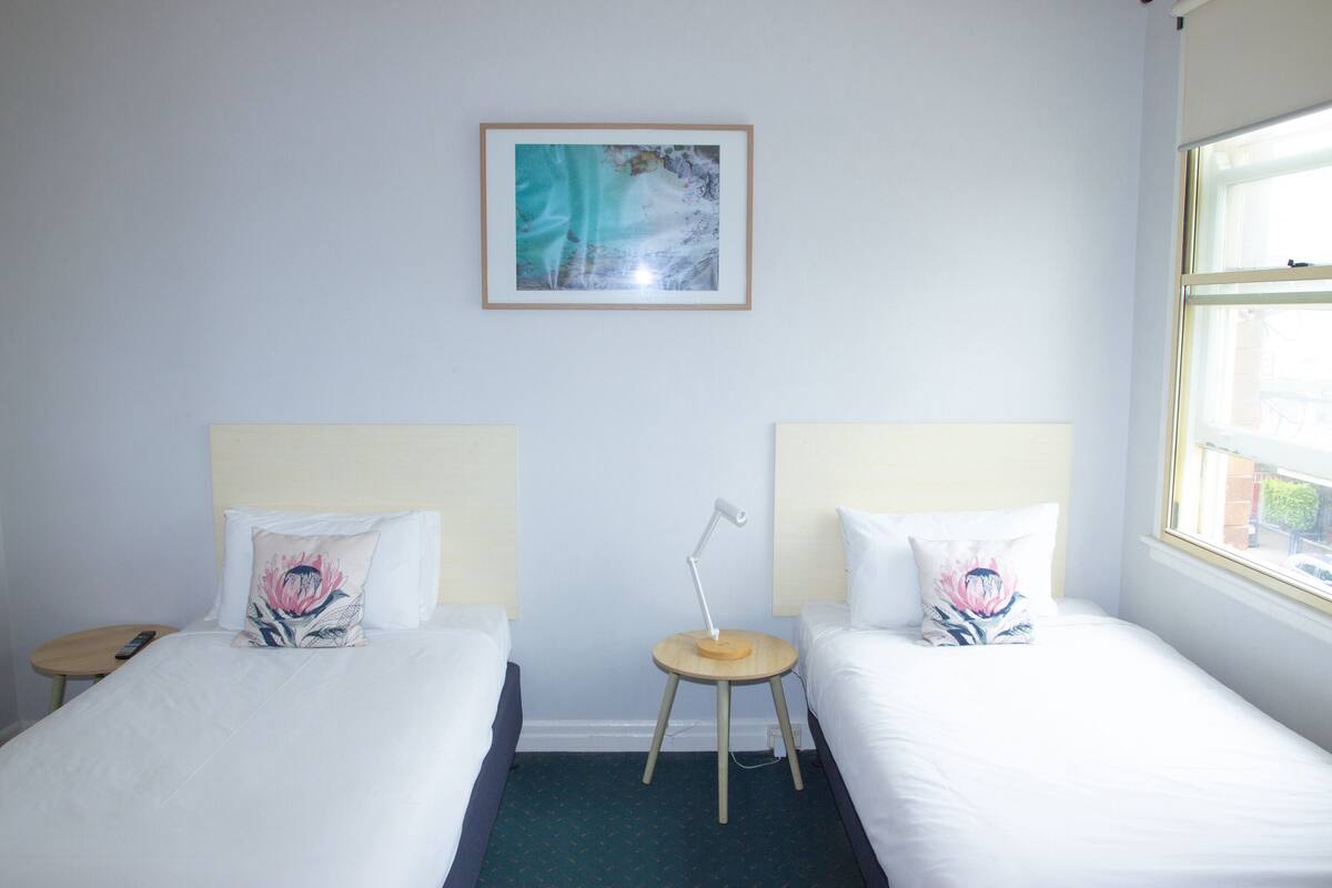 Quarto de hotel com duas camas de solteiro, cada uma com almofada e mesa de cabeceira. Há um quadro na parede e uma janela no lado direito.