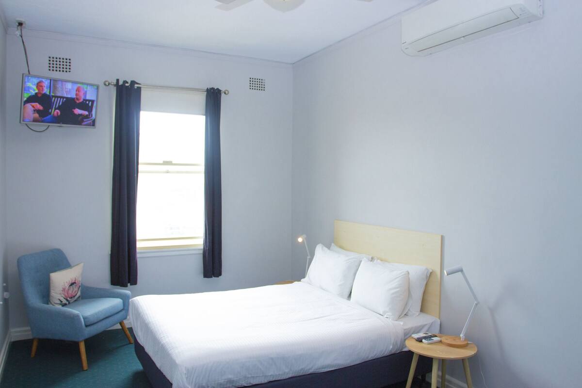 Quarto de hotel com cama, cadeira, televisão e janela com cortinas.