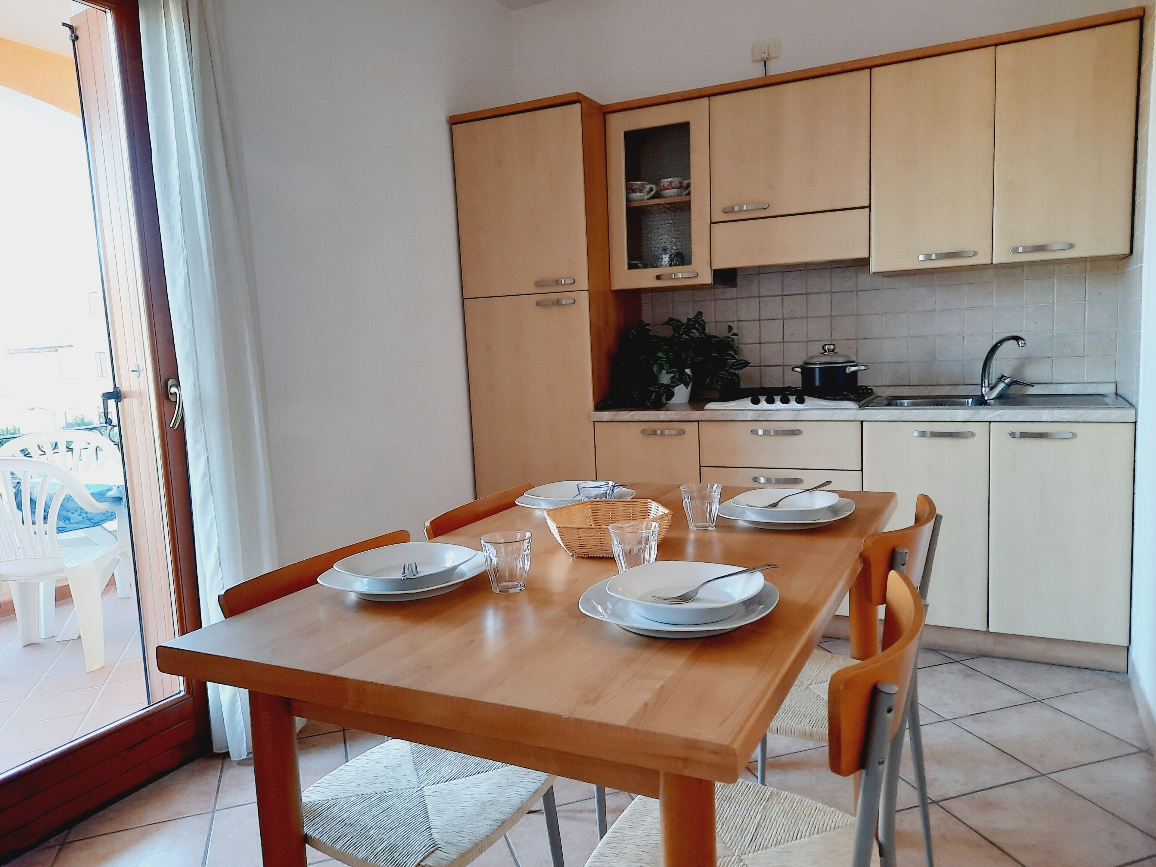 Cozinha com armários de madeira, uma mesa de jantar posta com pratos e copos, e vista para o exterior por uma porta de vidro.