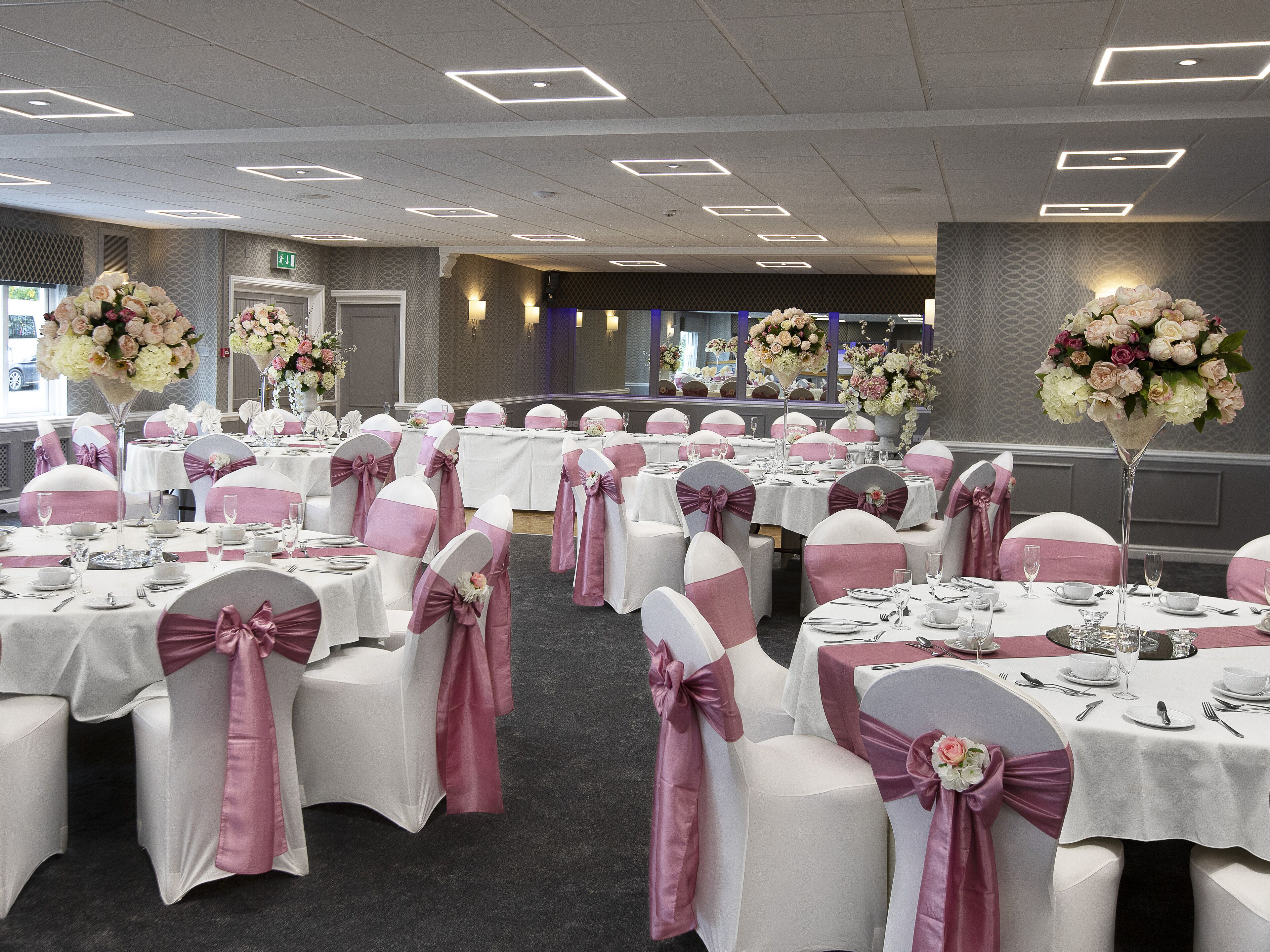 Salão de banquetes com mesas redondas, cada uma com toalhas brancas e fitas cor-de-rosa.