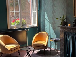 Quarto com duas poltronas, uma mesinha, um radiador, uma janela com vista para um muro de tijolos e flores vermelhas, e uma lareira com cortina.