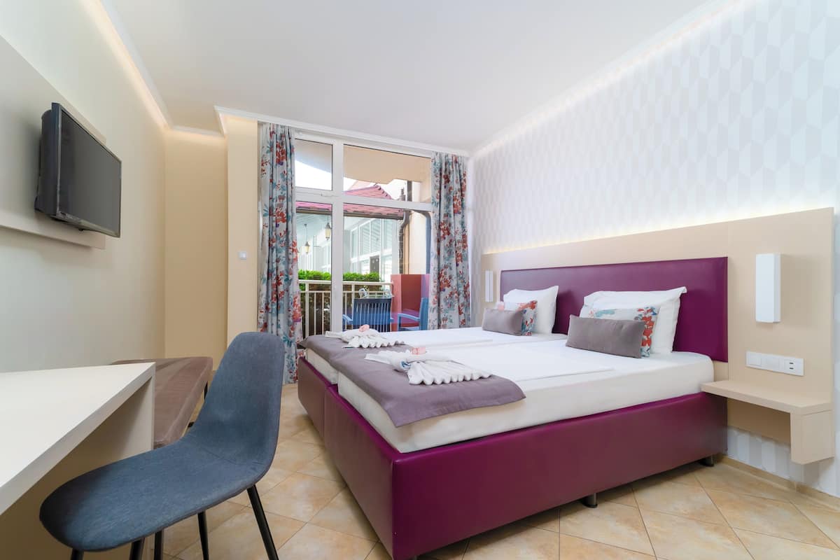 Quarto de hotel moderno com uma cama grande, uma escrivaninha e uma cadeira. Há uma TV na parede, e o quarto oferece vista para o exterior.