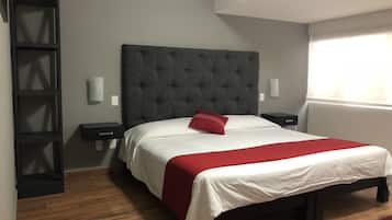 Quarto moderno com cabeceira estofada, piso de madeira, colcha vermelha e branca e mesa de cabeceira com luminária.