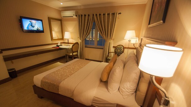 Quarto de hotel com cama, mesa de cabeceira, luminária, escrivaninha, cadeira, televisão e cortinas.