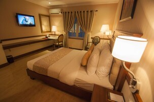 Quarto de hotel com cama, mesa de cabeceira, luminária, escrivaninha, cadeira, televisão e cortinas.
