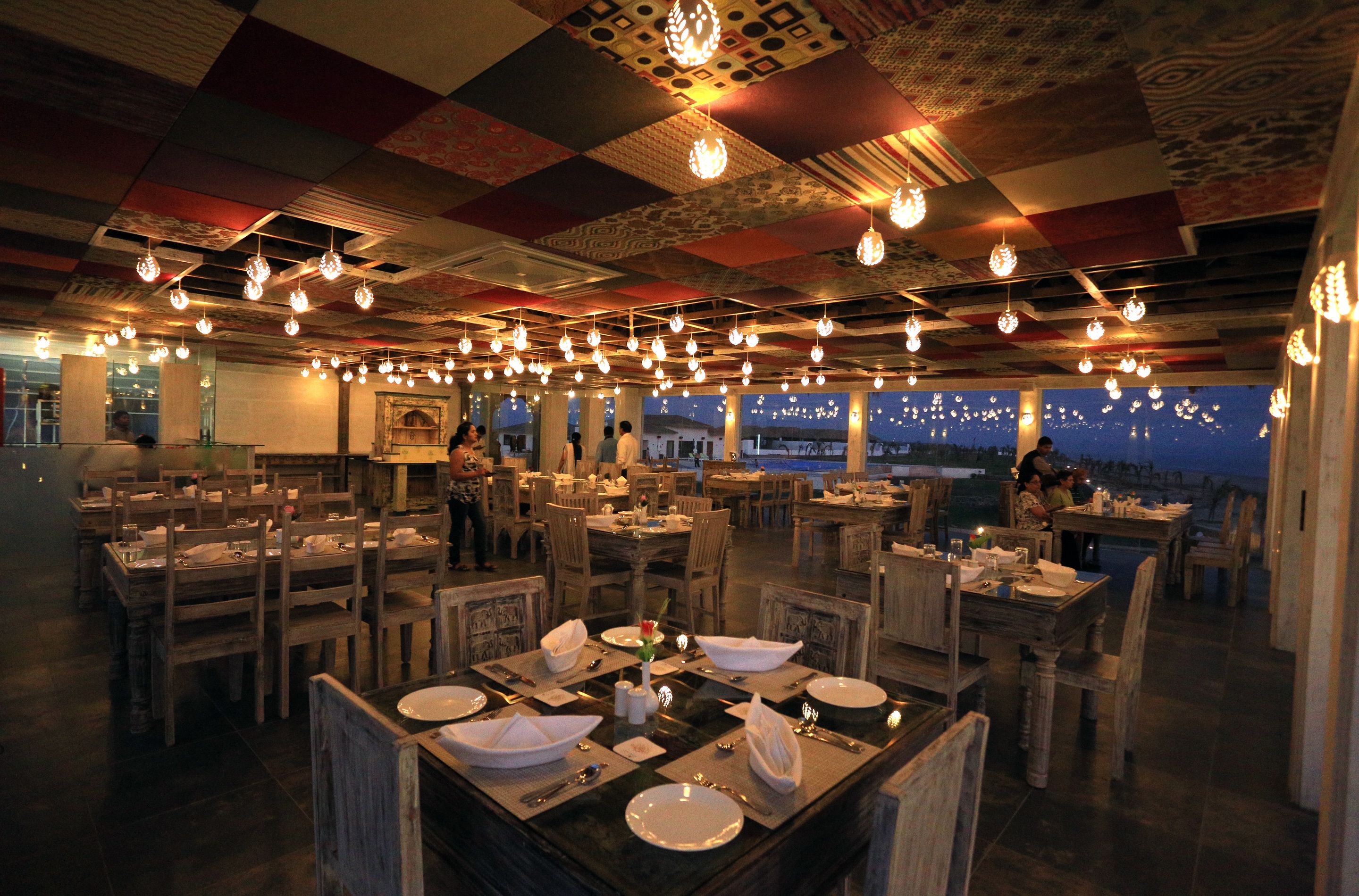 Restaurante com várias mesas preparadas para os hóspedes, com teto adornado por luminárias pendentes e vista para o exterior.