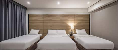 Quarto de hotel com três camas, cabeceira de madeira, mesa de cabeceira com abajur e uma janela grande com cortinas.