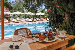 Mesa de jantar com comida e bebidas, com vista para uma piscina com espreguiçadeiras e guarda-sóis.