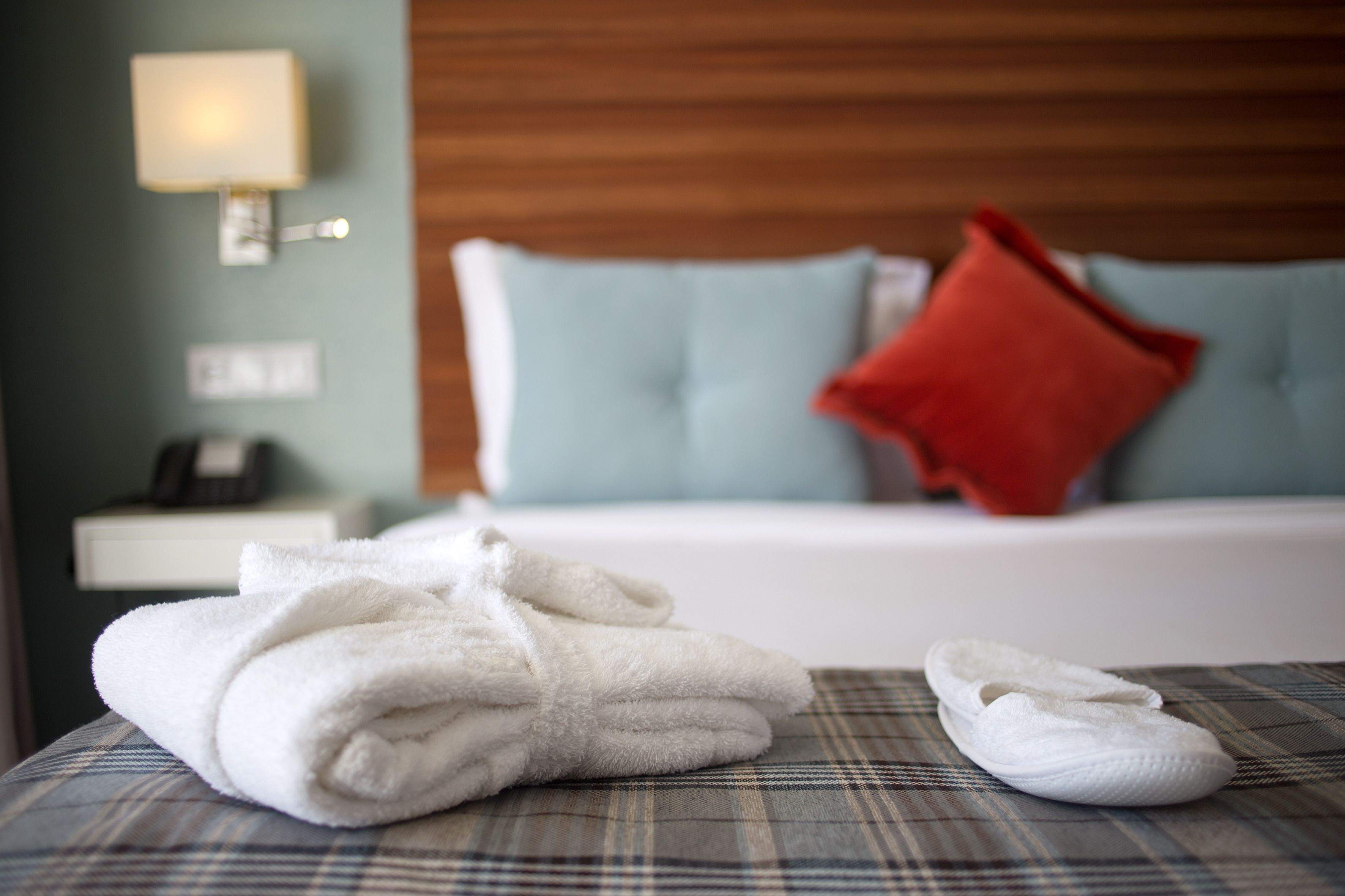 Uma cama bem arrumada com colcha xadrez, um travesseiro vermelho e uma toalha e chinelos brancos sobre a cama.