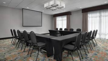 Sala de conferências com uma mesa comprida, cadeiras, tela de projetor e grandes janelas com cortinas.