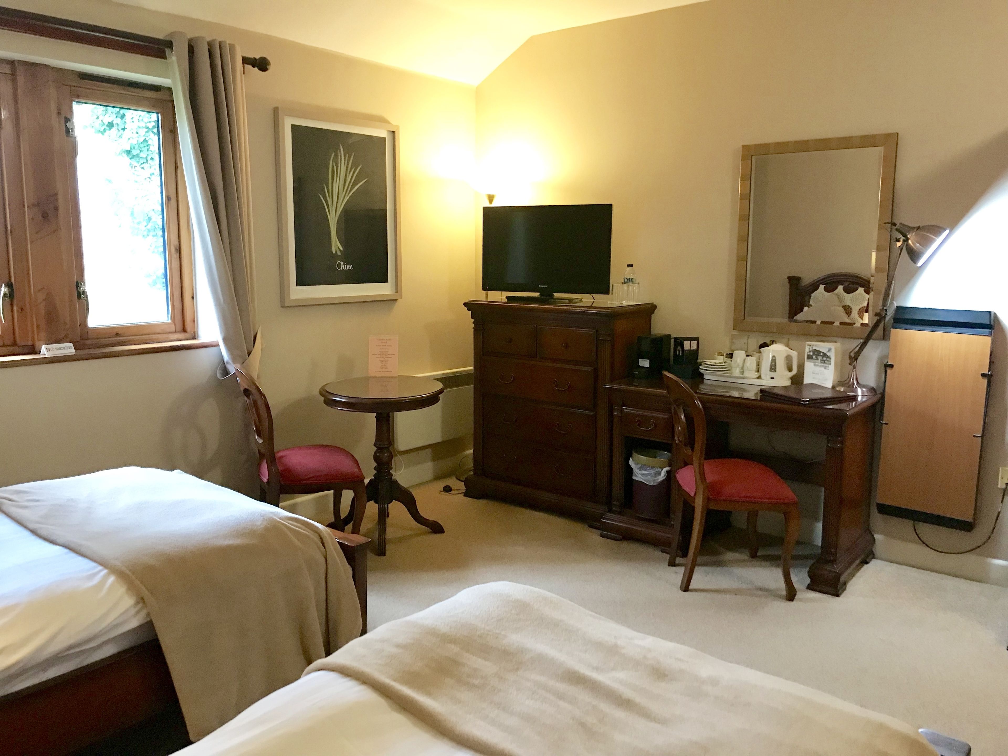 Quarto de hotel com duas camas, uma escrivaninha com computador, uma cadeira, cômoda, espelho e janela com cortinas.
