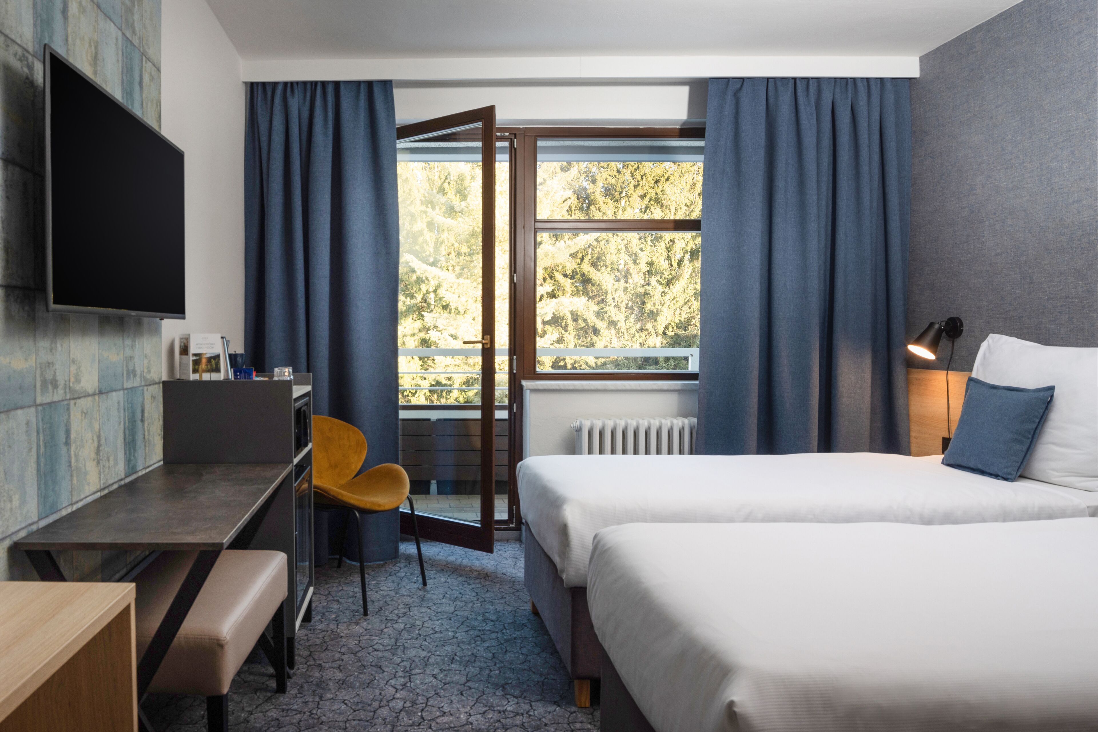 Quarto de hotel moderno com uma cama, escrivaninha, cadeira, televisão e janela com cortinas.