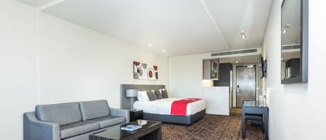 Quarto de hotel moderno com sofá cinza, cama com detalhe em vermelho, mesa de centro e piso coberto por carpete.