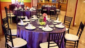 Uma sala de banquetes com mesas redondas cobertas por toalhas roxas, rodeadas por cadeiras pretas com almofadas brancas.
