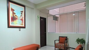 Uma área de espera com um banco, duas cadeiras com almofadas laranja, um quadro emoldurado e uma porta com placa.