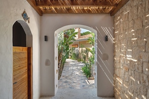 Corredor com parede de pedra à direita, porta de madeira à esquerda e caminho de azulejos que leva a um edifício com telhado em azulejos.