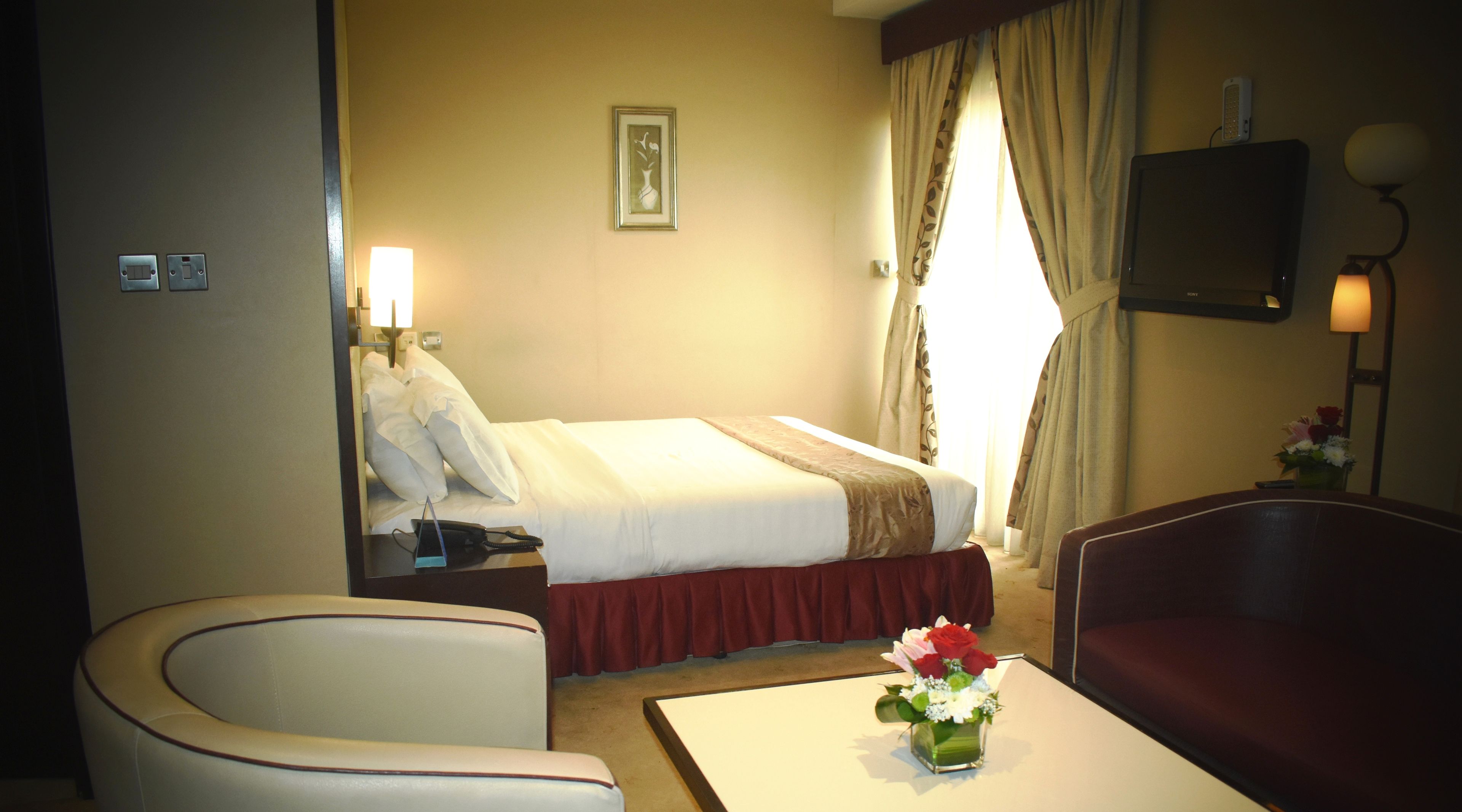 Quarto de hotel com cama, poltrona, mesa, televisão e um vaso com flores.