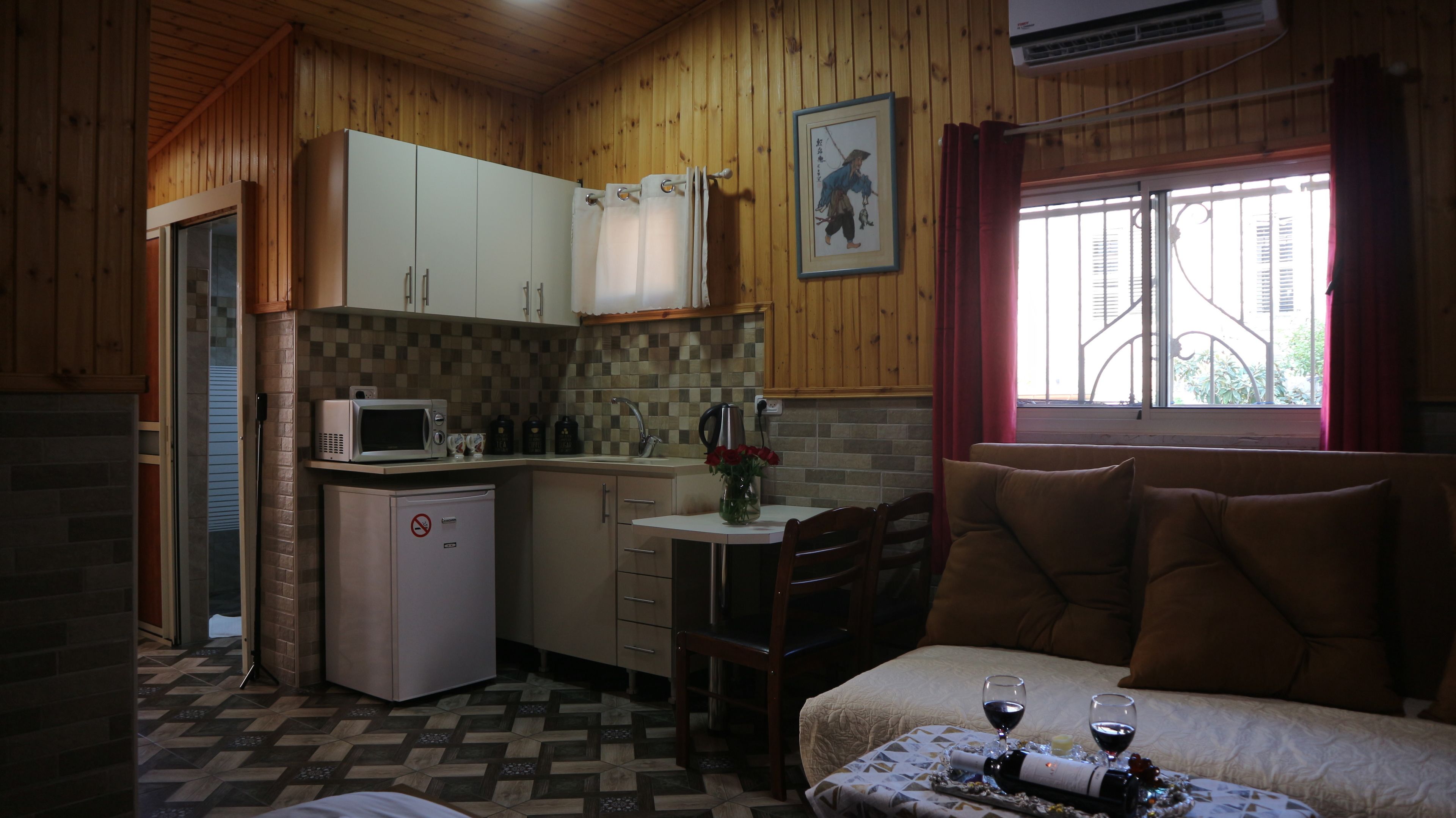 Uma cozinha compacta com micro-ondas, pia e armários. Há uma área de refeições com uma mesa posta para dois, um sofá e uma janela com cortinas.