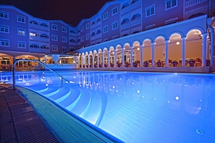Uma área de piscina com um elemento aquático, um espaço coberto com mesas e cadeiras, e um edifício com várias varandas.