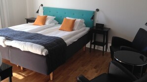 Uma cama bem arrumada com cabeceira verde-água, dois travesseiros e um cobertor. Há duas mesas de cabeceira, uma com abajur, e uma pequena mesa redonda com uma cadeira.