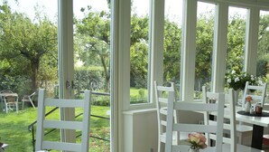 Espaço de refeições com cadeiras e mesas brancas, amplas janelas com vista para um jardim com árvores e uma bicicleta.