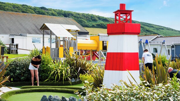 Um jardim ajardinado com um farol vermelho e branco, um pequeno lago e um edifício com telhado inclinado ao fundo.