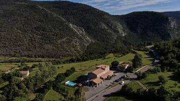Um edifício grande com vários quartos, piscina e estacionamento, cercado por campos verdes e montanhas.