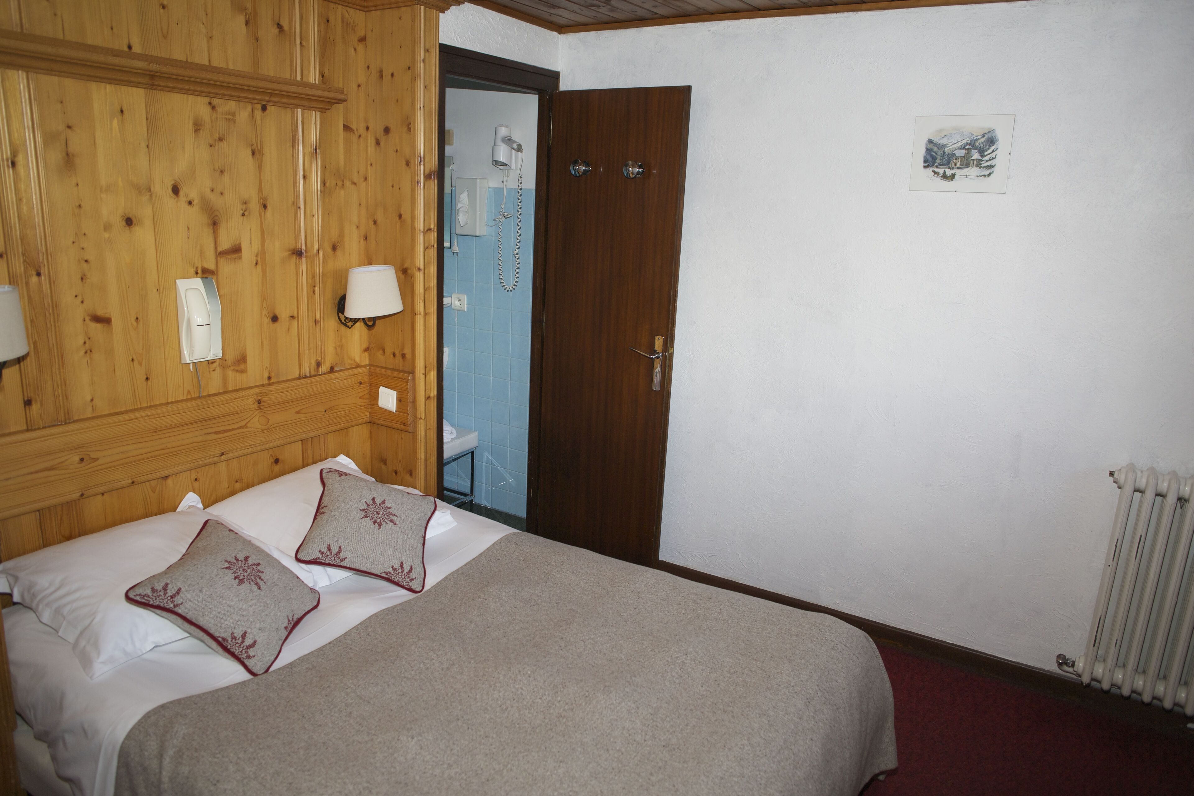 Quarto com cabeceira de madeira, cama com roupa de cama branca e almofadas decorativas, porta de madeira e um pequeno quadro na parede.