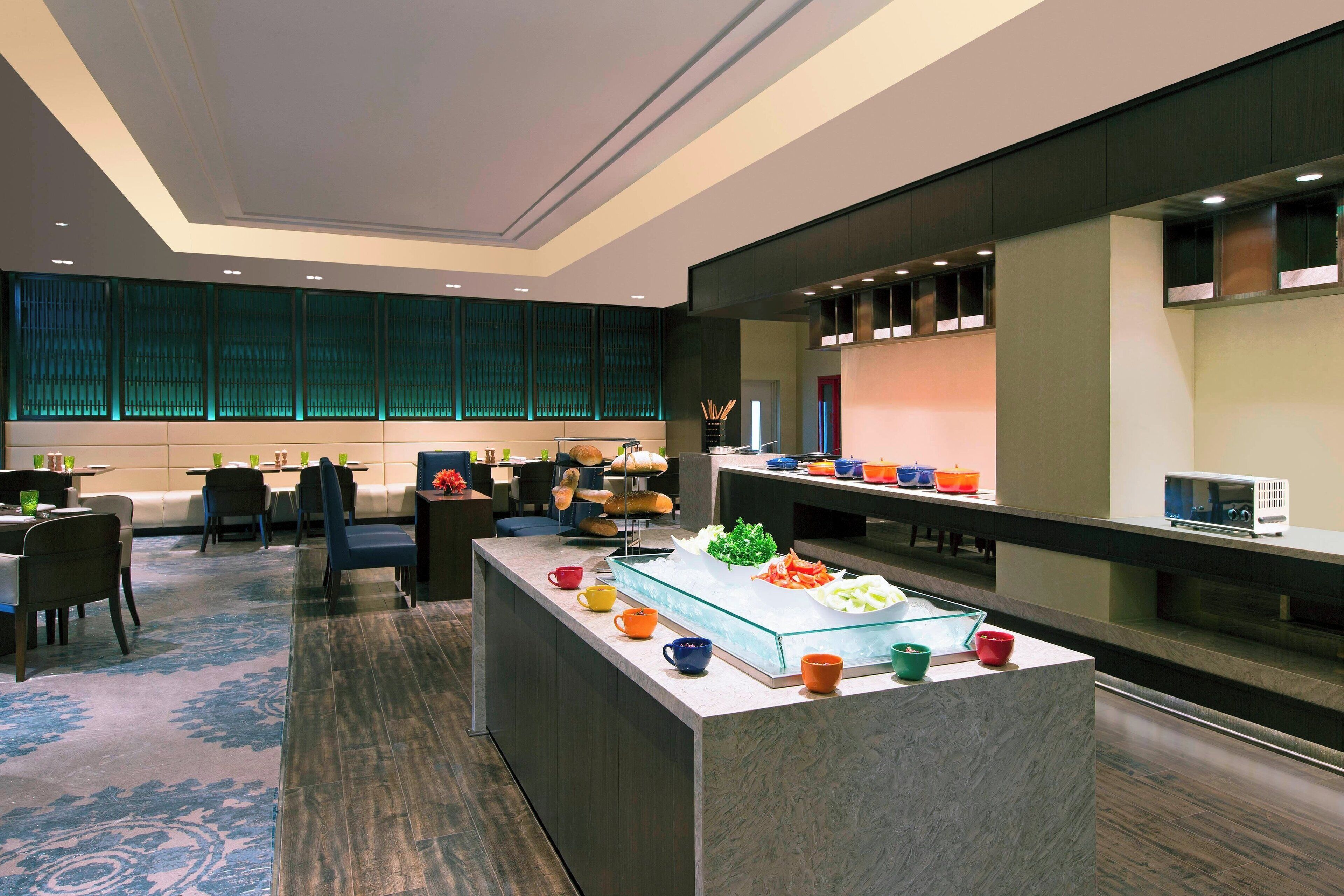 Um saguão de hotel moderno com um buffet central, áreas de estar e uma grande janela com vista.