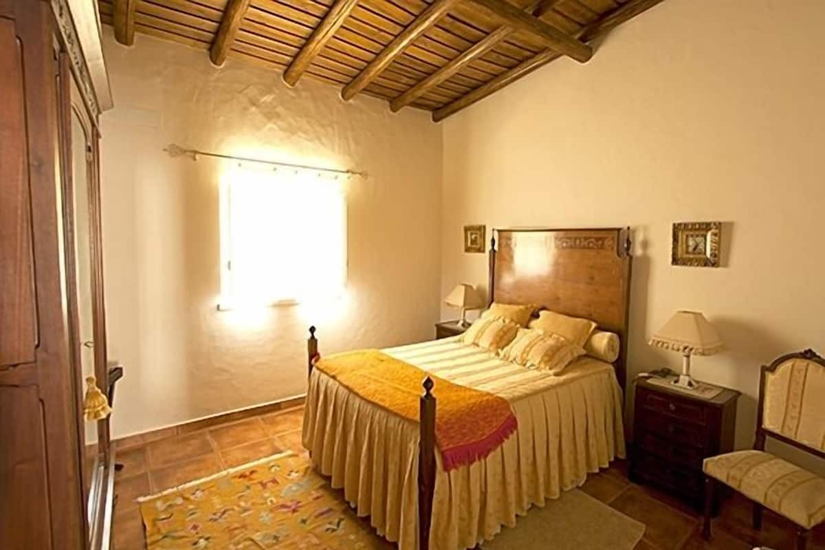 Um quarto com teto de madeira, uma cama com colcha amarela, uma mesa de cabeceira, uma cadeira e uma janela com cortinas.