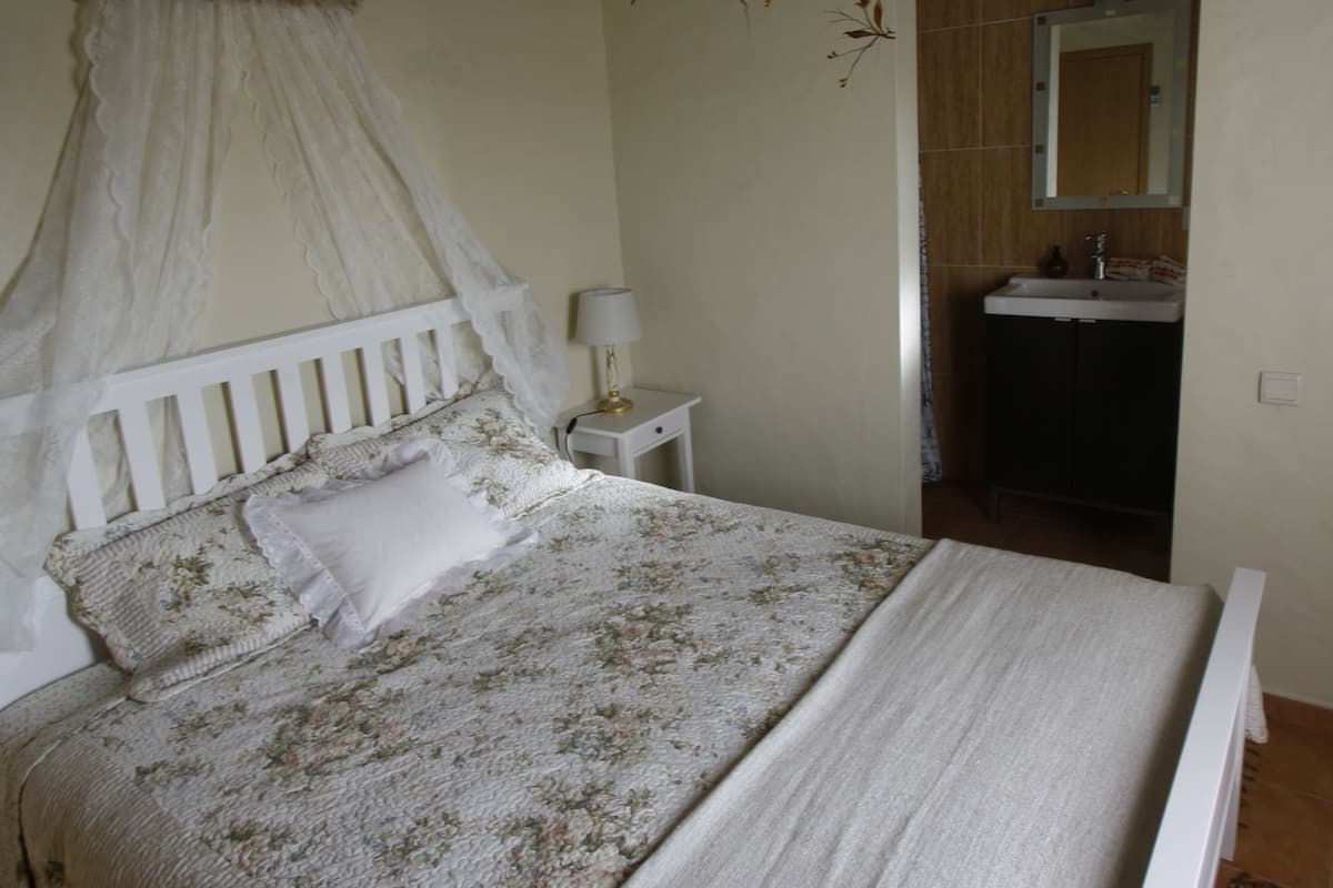 Quarto com cama de dossel, mesa de cabeceira com abajur e banheiro com pia e espelho.