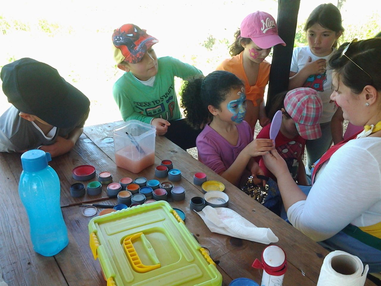 Um grupo de pessoas participando de uma atividade de pintura em uma mesa de madeira, com diversos potes de tinta e pincéis.
