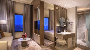 Quarto de hotel com cama, área de estar com mesa de centro, banheiro com espelho e pia, e vista da cidade à noite.