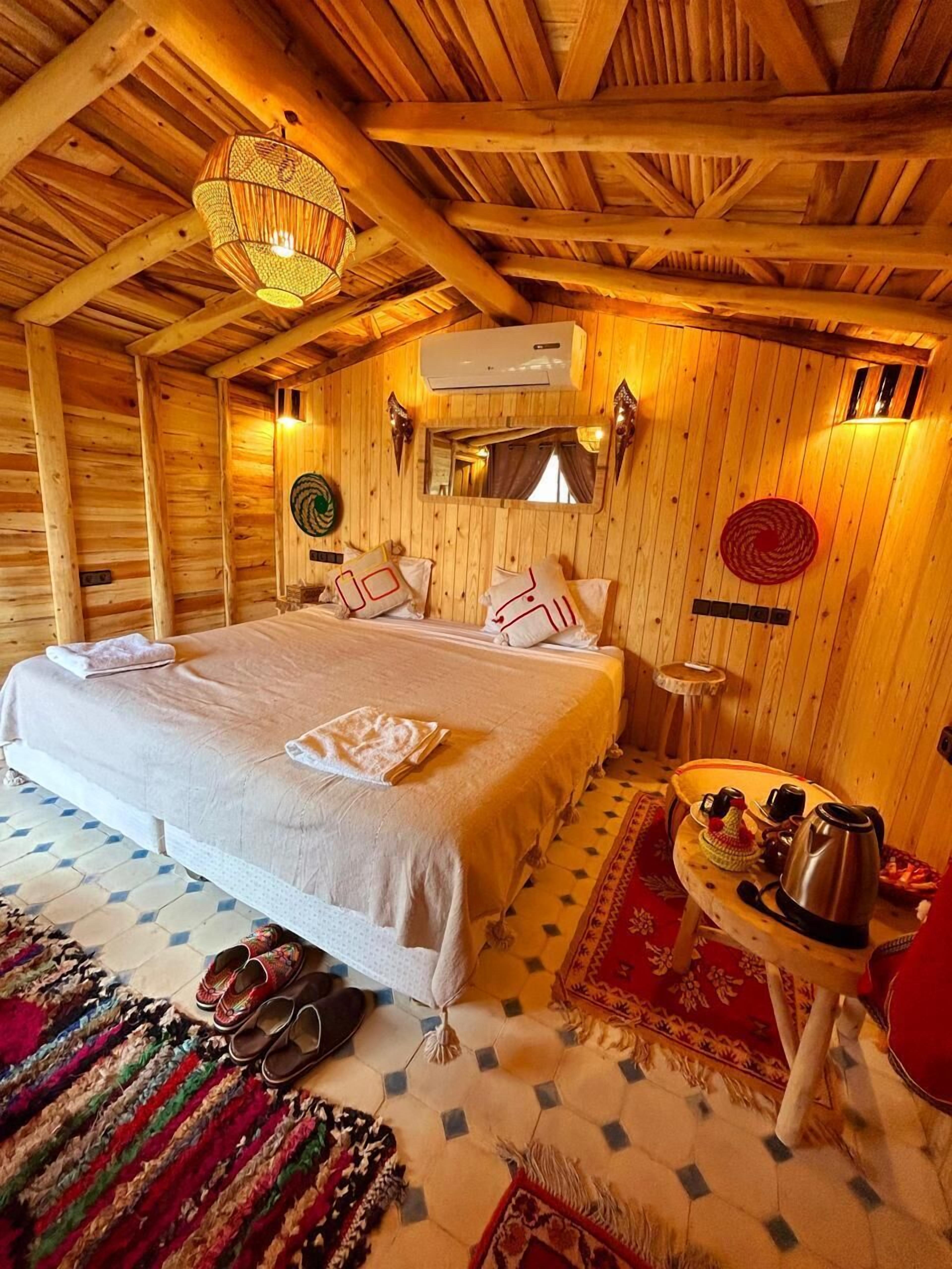 Um quarto de madeira com uma cama, uma mesinha com chaleira e um tapete.