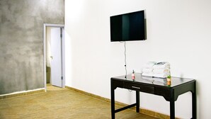 Quarto com televisão de tela plana na parede, uma mesinha com toalhas dobradas e um vaso de planta, e uma porta que dá para outro cômodo.