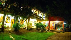 Hotel bem iluminado com entrada pavimentada, área gramada com pedras e um prédio vermelho com entrada visível.