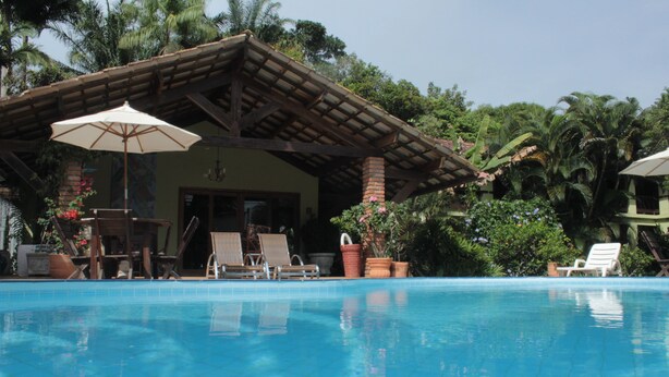 Piscina azul cristalina com espreguiçadeiras e guarda-sóis, cercada por vegetação exuberante e um pavilhão de madeira.