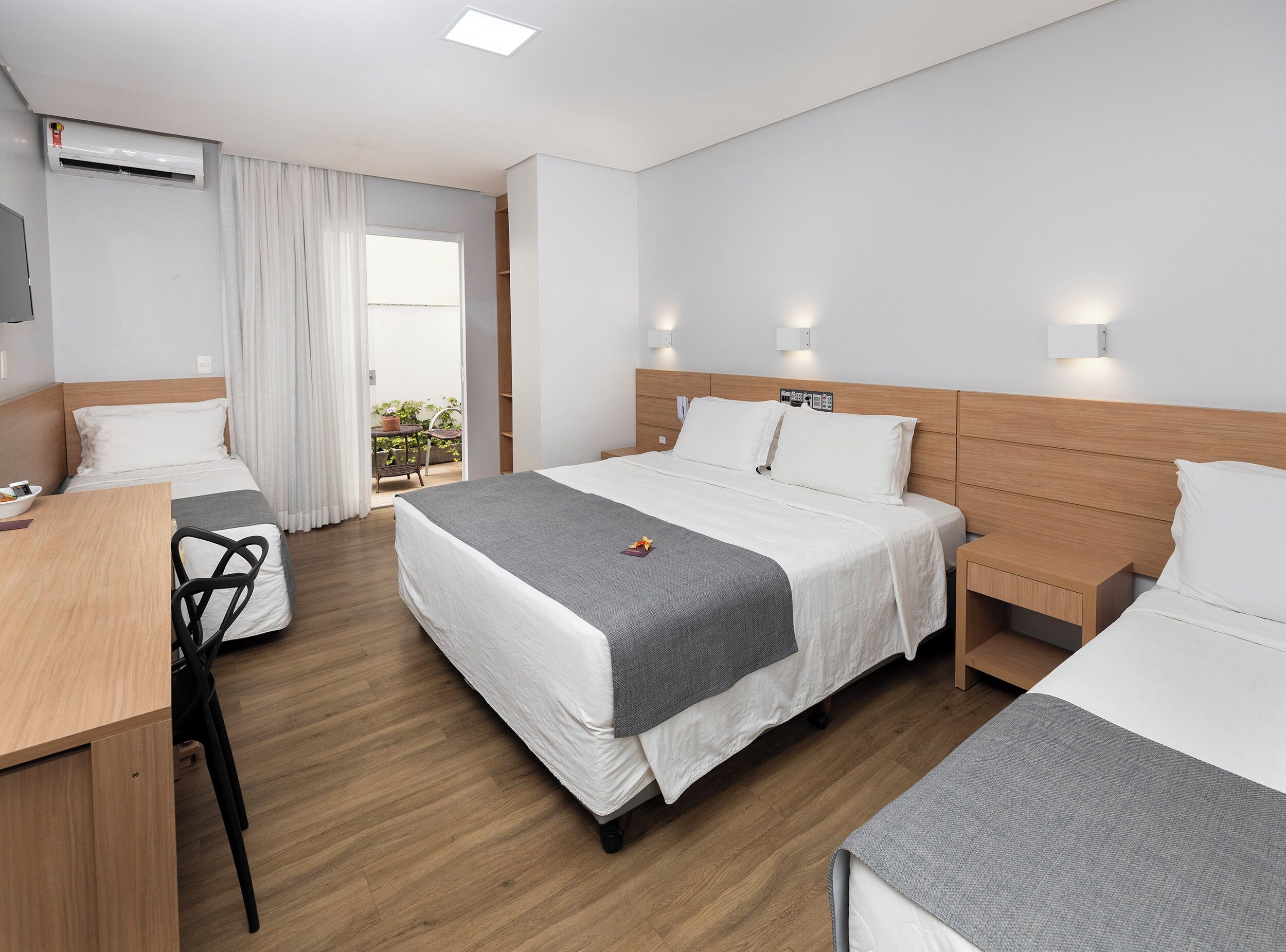 Quarto de hotel com uma cama grande, uma escrivaninha e uma cadeira. Há uma janela com cortinas, um ar condicionado fixado na parede e uma pequena mesa com um vaso de planta.