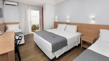 Quarto de hotel com uma cama grande, uma escrivaninha e uma cadeira. Há uma janela com cortinas, um ar condicionado fixado na parede e uma pequena mesa com um vaso de planta.