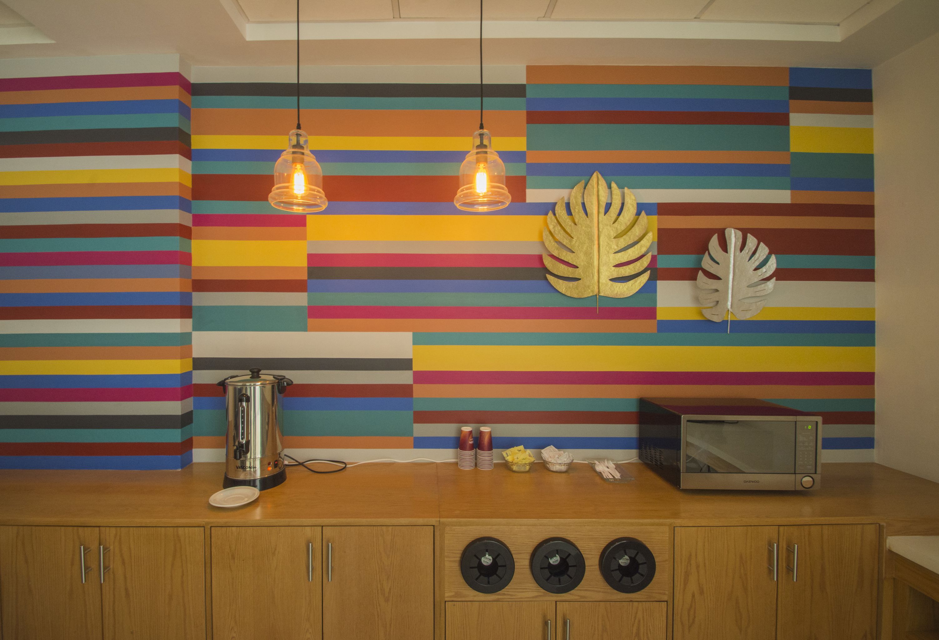 Cozinha com parede listrada e colorida, uma cafeteira, uma torradeira e duas luminárias pendentes.