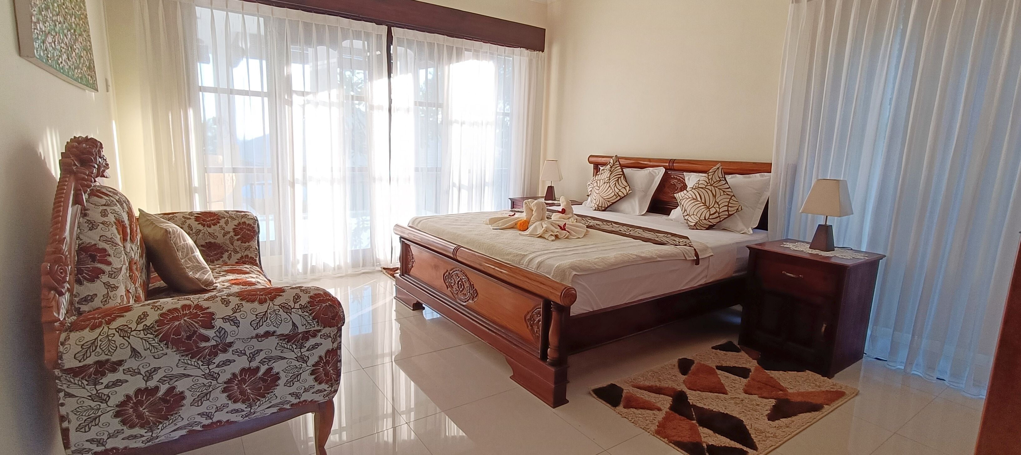 Quarto com cama de madeira, poltrona floral, mesa de cabeceira com abajur e uma grande janela com cortinas leves.